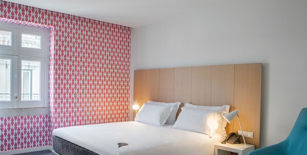 Quarto de hotel com cama, mesa de cabeceira com luminária, cadeira e janela com vista.