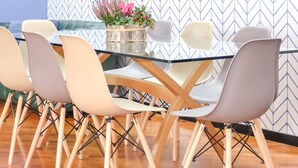 Uma sala de jantar com uma mesa de vidro rodeada por cadeiras modernas, um sofá azul e uma parede estampada.