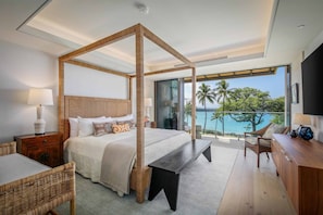 Quarto moderno com cama de dossel, mesa de cabeceira de madeira, cadeira de vime, escrivaninha de madeira e uma grande janela com vista para o mar.