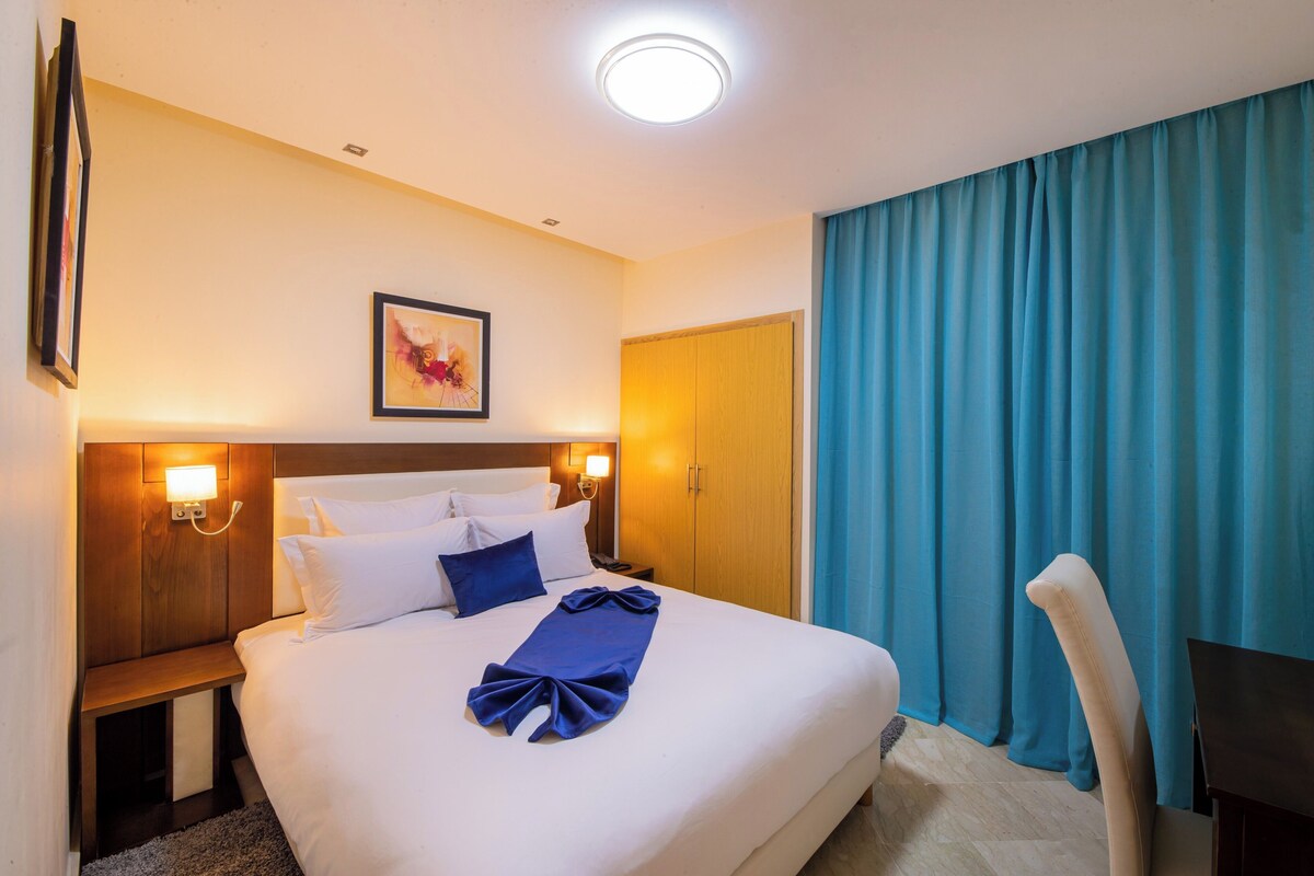 Quarto de hotel com cama, mesas de cabeceira, escrivaninha, cadeira, cortina e um quadro na parede.