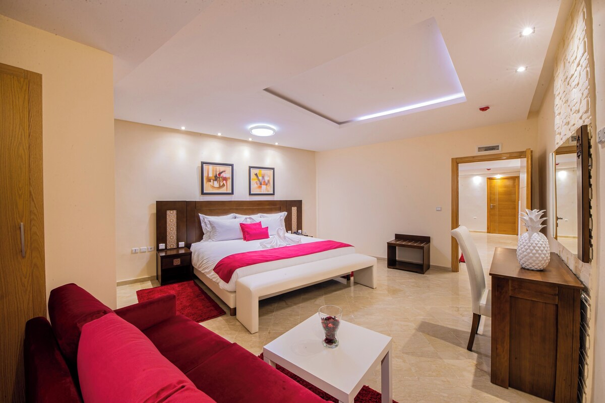 Quarto de hotel moderno com uma cama, um sofá vermelho, uma pequena mesa branca e um espelho.