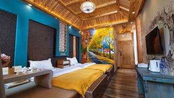 Quarto de hotel com teto de madeira, cama com cobertor amarelo, escrivaninha com chaleira e televisão na parede.