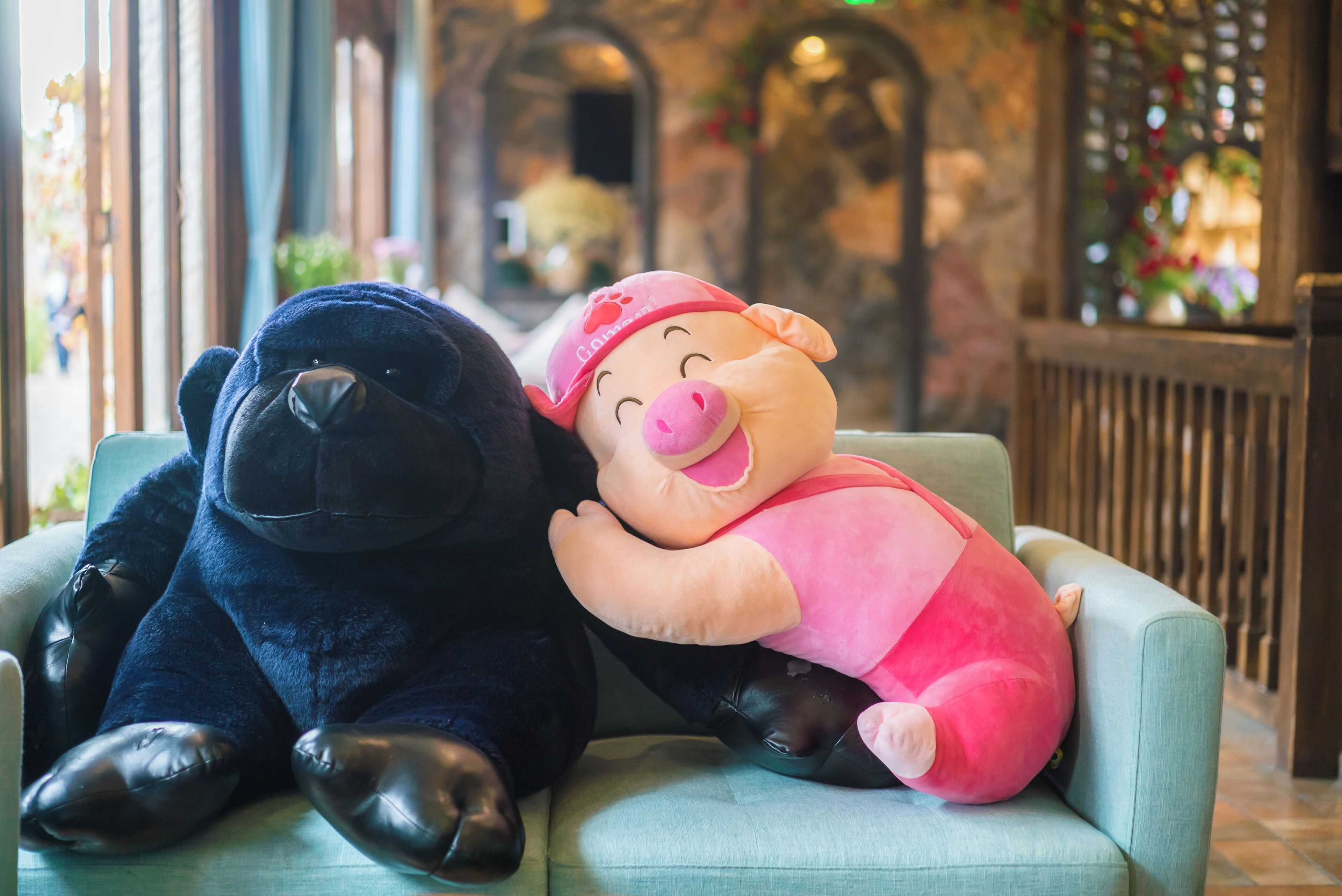 Dois brinquedos de pelúcia, um urso preto e um porquinho rosa, sentados num sofá verde claro num quarto com paredes de pedra e um corrimão de madeira.