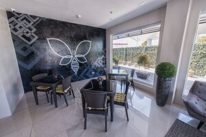 Uma zona de refeições moderna com mesa de vidro e cadeiras de vime, uma grande parede decorativa com padrões geométricos e vista para o exterior através de grandes janelas.