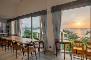 Uma área de refeições com uma grande mesa de madeira e cadeiras, janelas amplas com cortinas, e vista para o pôr do sol sobre a água.