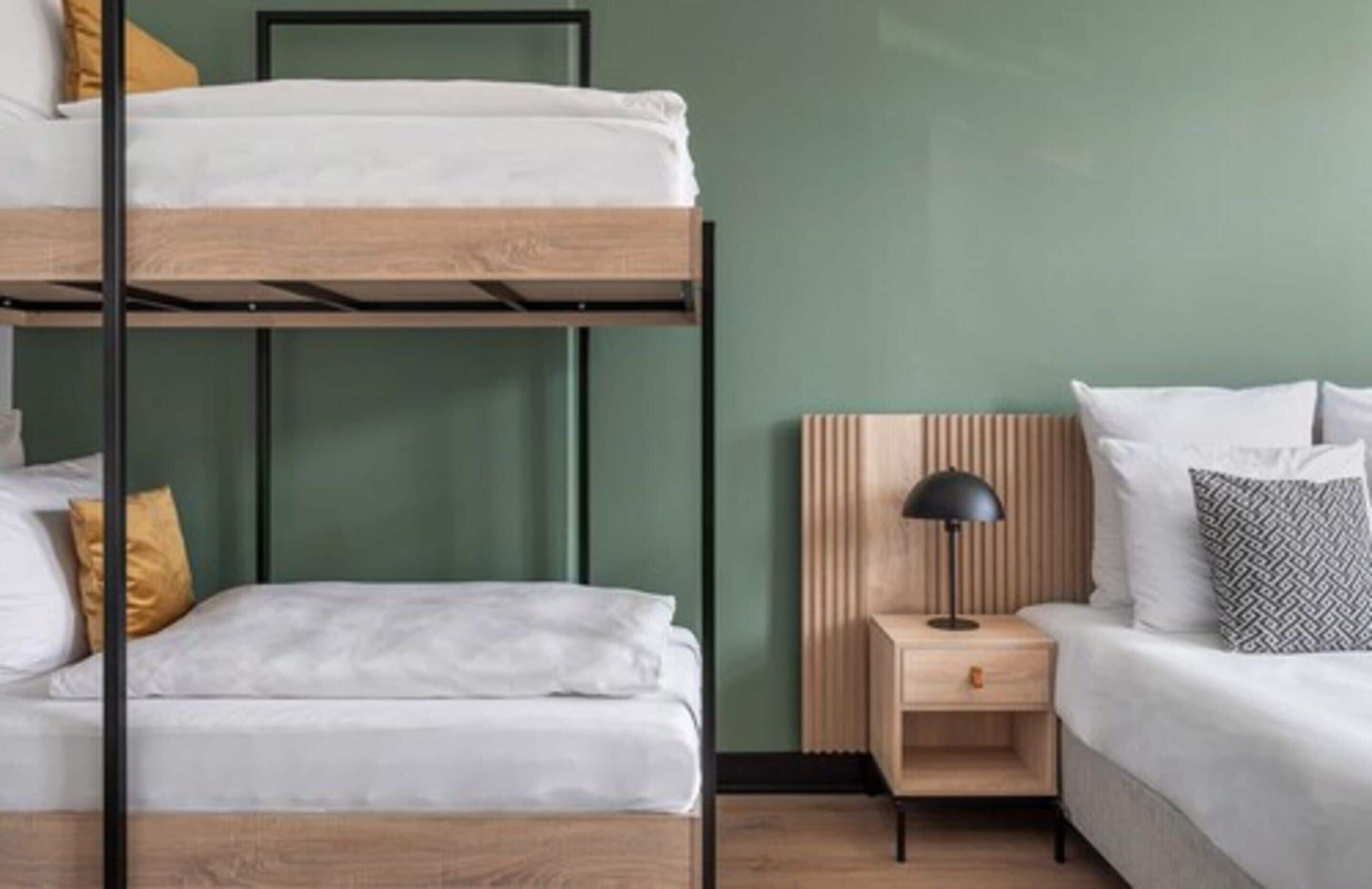 Quarto com beliche, cabeceira de madeira, mesa de cabeceira com abajur e uma cama de solteiro com almofadas.