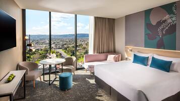 Quarto de hotel com uma cama grande, área de estar com uma mesa e vista para a paisagem urbana.