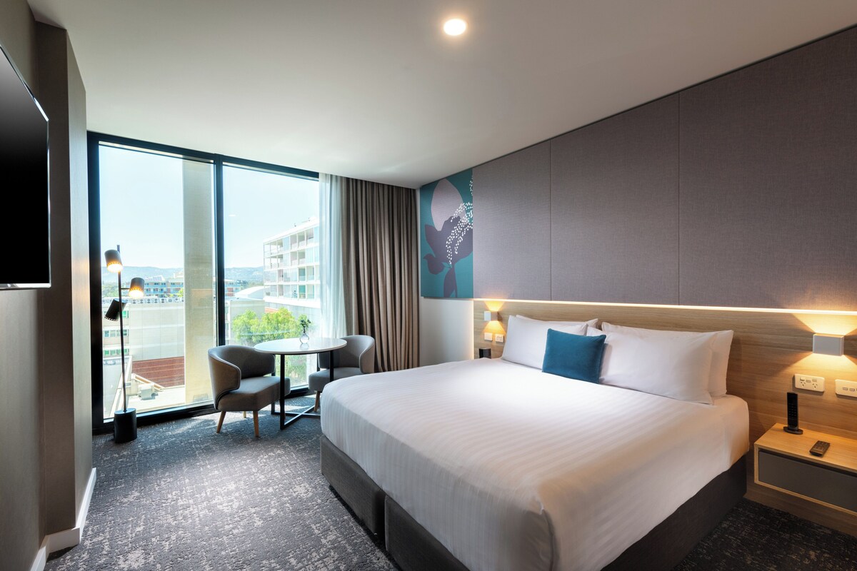 Quarto de hotel com uma cama grande, uma escrivaninha, uma cadeira, uma televisão e vista para a cidade.