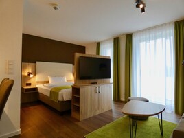 Quarto de hotel com uma cama, televisão fixada num armário de madeira, uma mesinha e um tapete verde.