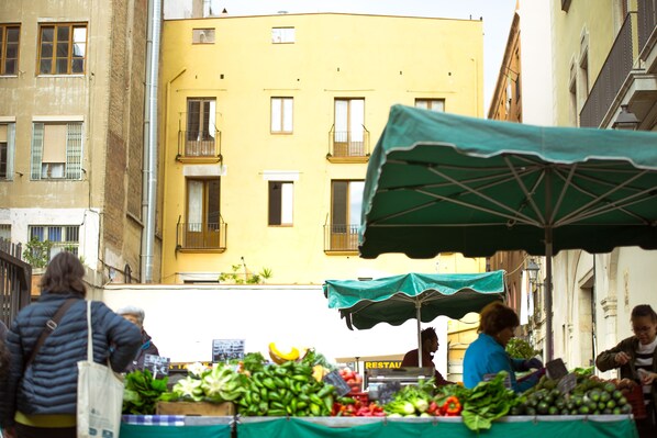 Mercado de rua com produtos frescos sob toldos verdes, edifícios ao fundo e pessoas fazendo compras.