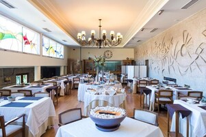 Uma ampla sala de jantar com várias mesas postas para refeição, um grande candelabro e um mural decorativo na parede.