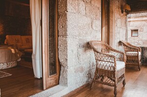 Quarto com parede de pedra, cadeiras de vime, piso de madeira e cama com colcha branca.