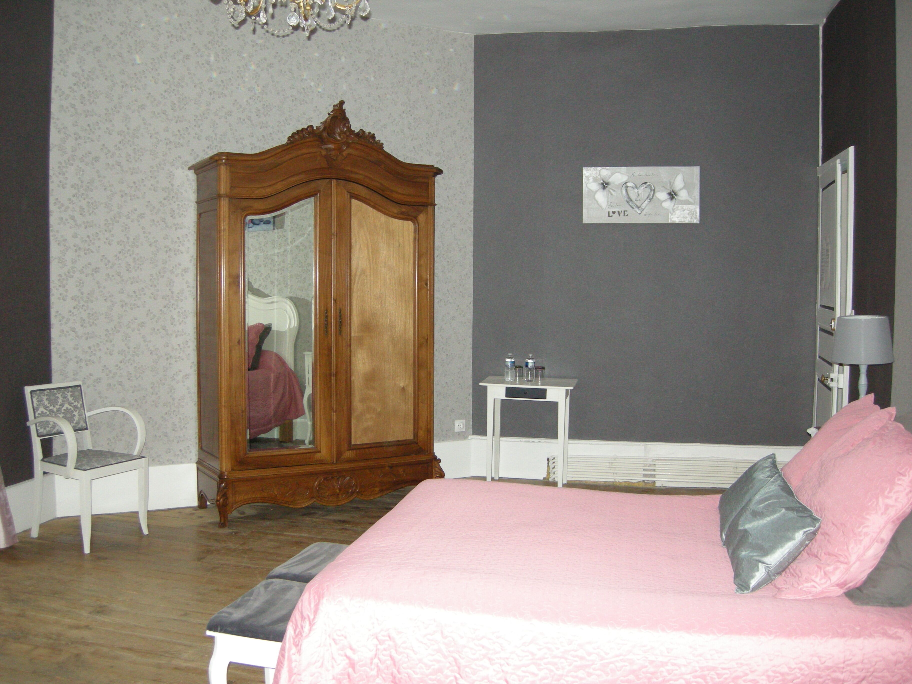 Um quarto com um guarda-roupa de madeira grande, uma cama com roupa de cama rosa, uma mesinha de cabeceira pequena e um espelho.