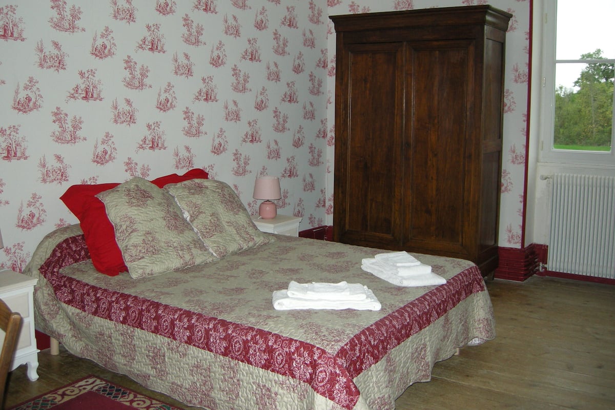 Quarto com cama, almofadas, mesa de cabeceira, guarda-roupa de madeira e janela.