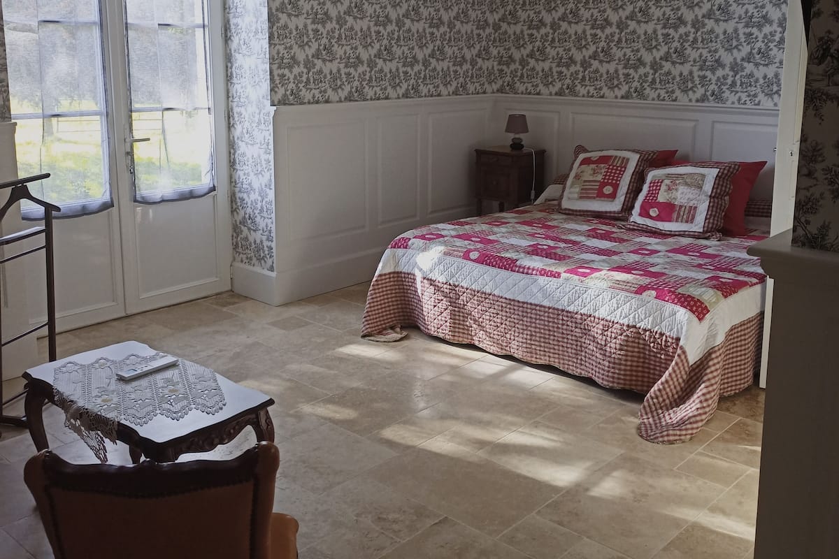 Quarto com uma janela grande, uma cama com colcha estampada, uma mesinha com toalha de renda e uma cadeira.