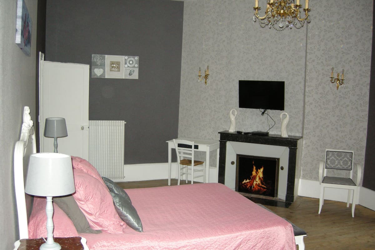 Quarto com cama, mesa de cabeceira com abajur, poltrona, lareira e televisão fixada na parede.