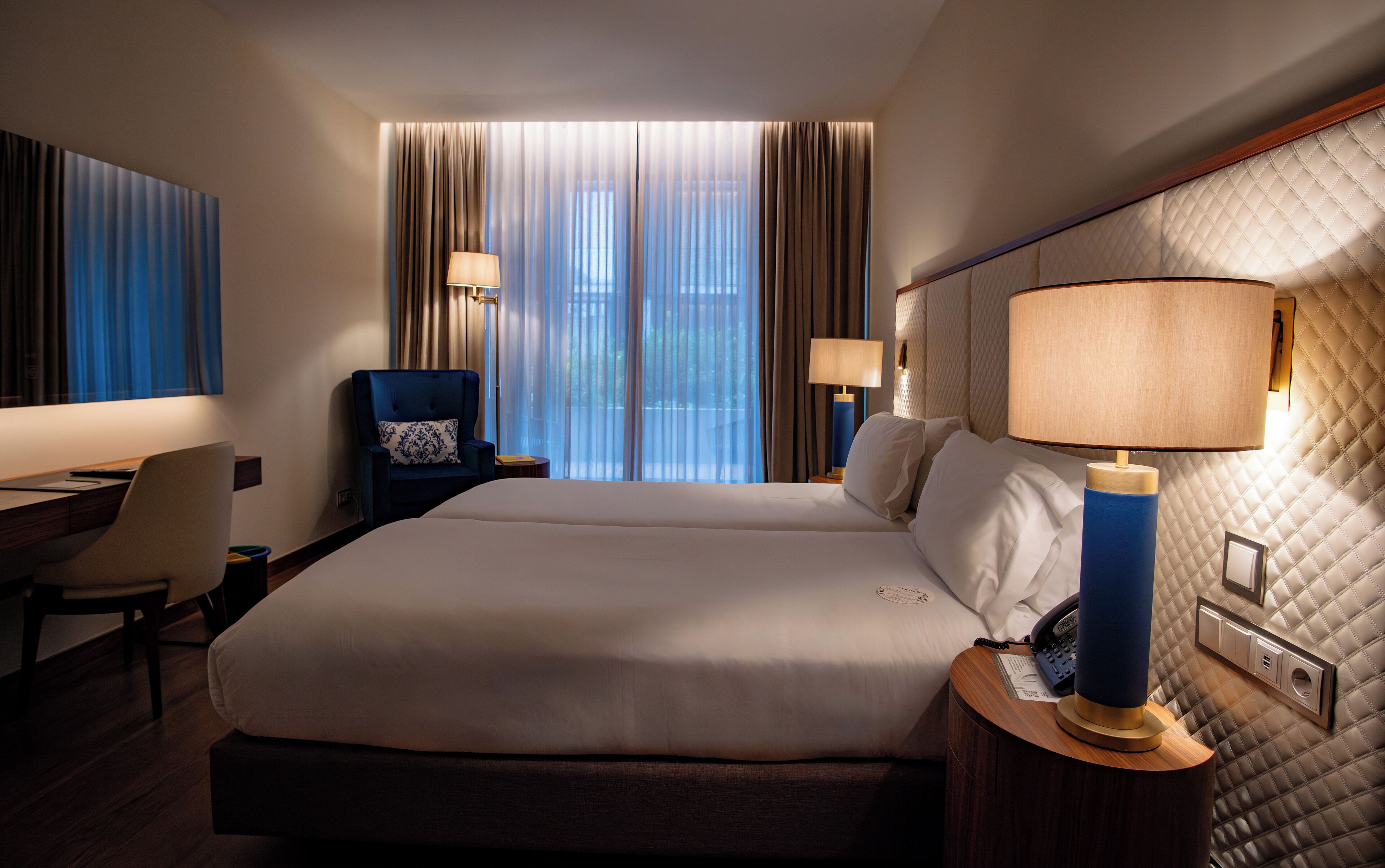 Quarto de hotel com uma cama grande, uma mesa, uma cadeira e duas luminárias de cabeceira.