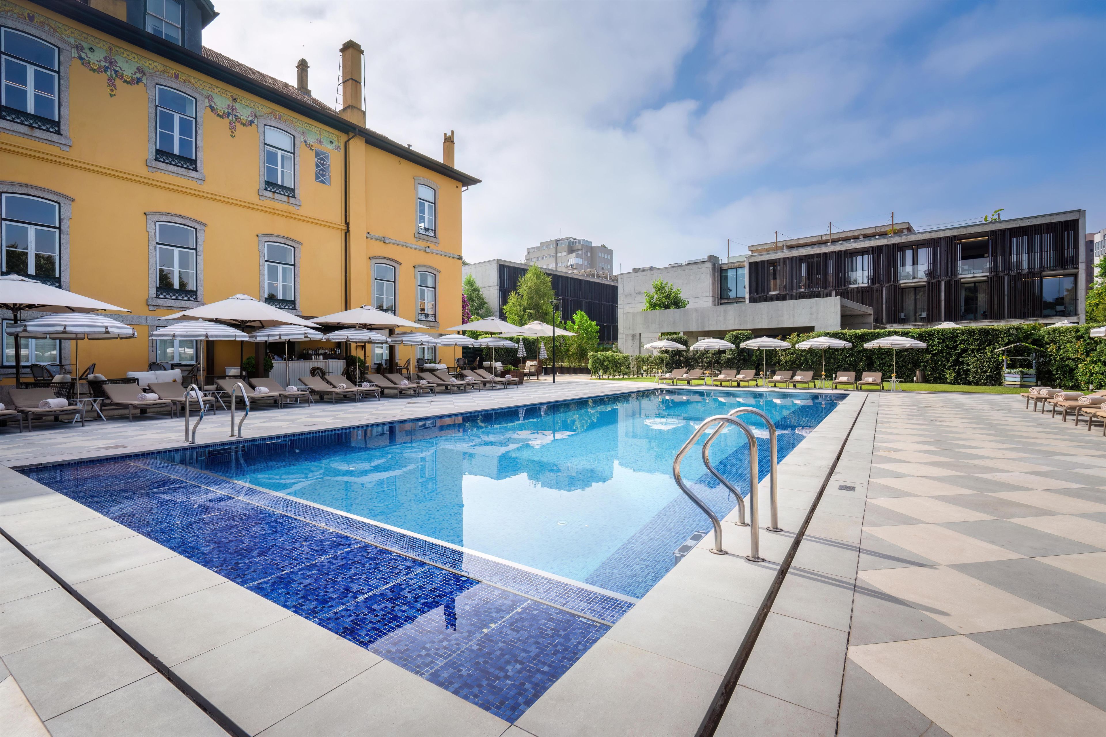 Piscina com espreguiçadeiras e guarda-sóis, rodeada por edifícios.