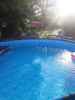 Piscina azul límpida com borda em azulejo, rodeada por vegetação e um guarda-sol vermelho.