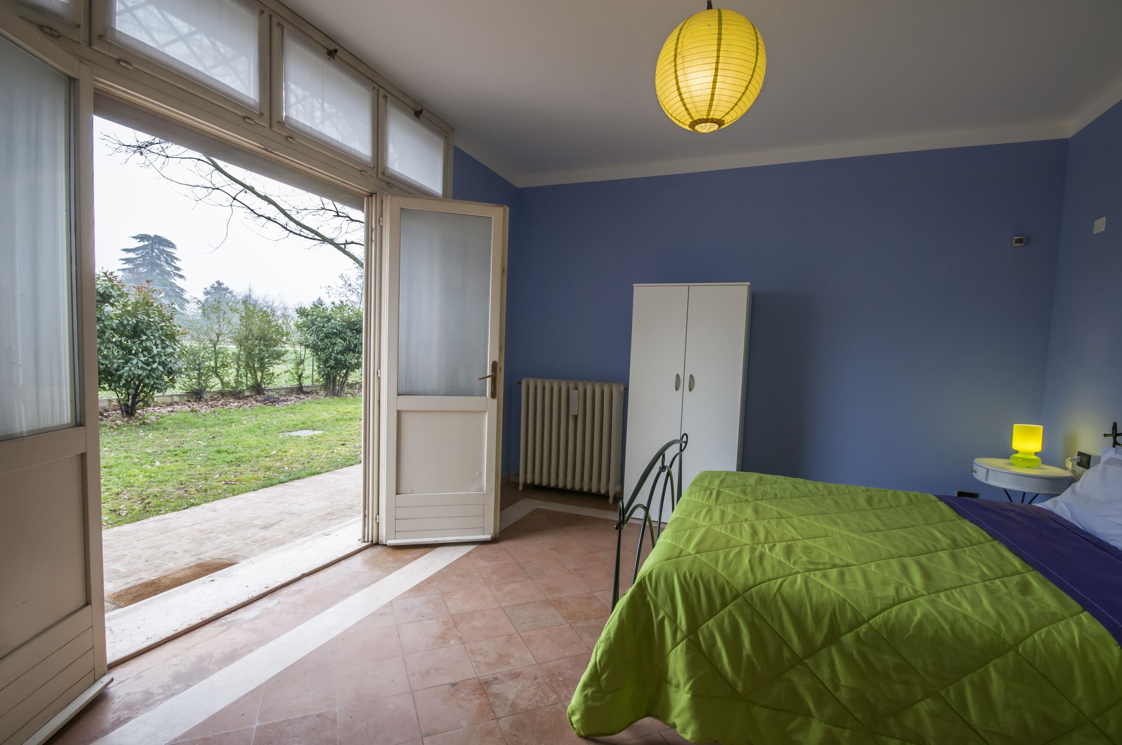 Quarto com colcha verde, guarda-roupa branco e uma pequena mesa de cabeceira com abajur.