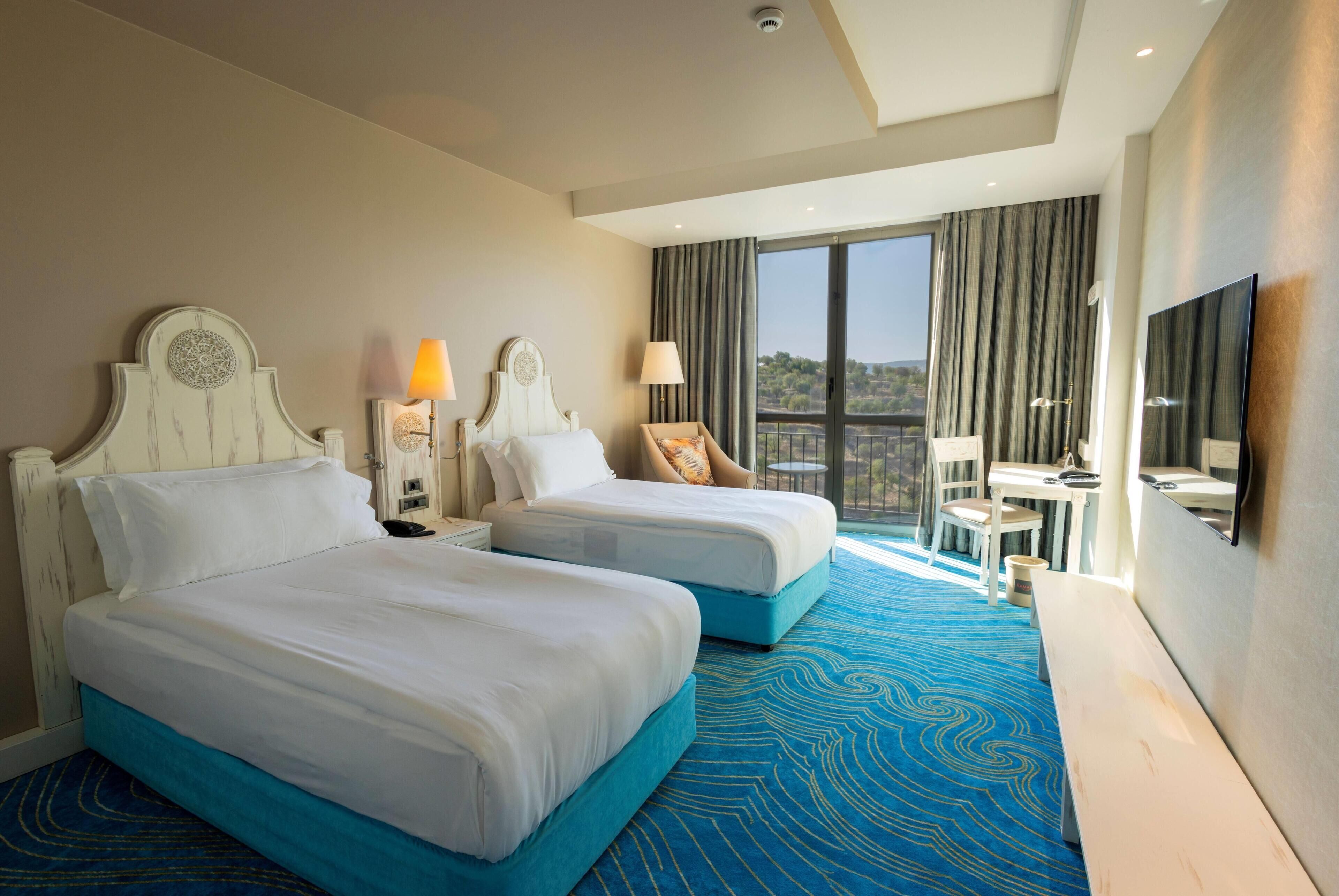 Quarto de hotel com duas camas, uma escrivaninha, uma cadeira e uma janela grande com vista.