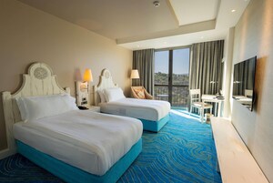 Quarto de hotel com duas camas, uma escrivaninha, uma cadeira e uma janela grande com vista.