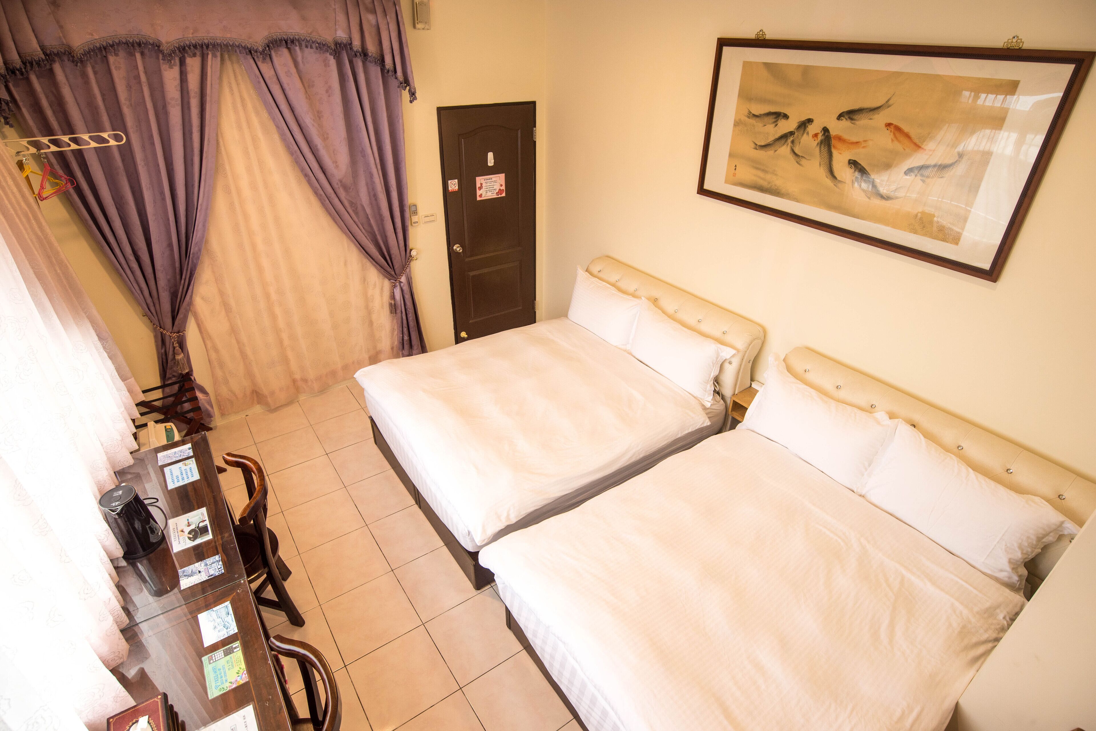 Quarto de hotel com duas camas, uma porta, um quadro emoldurado e uma mesa com chaleira e papéis.