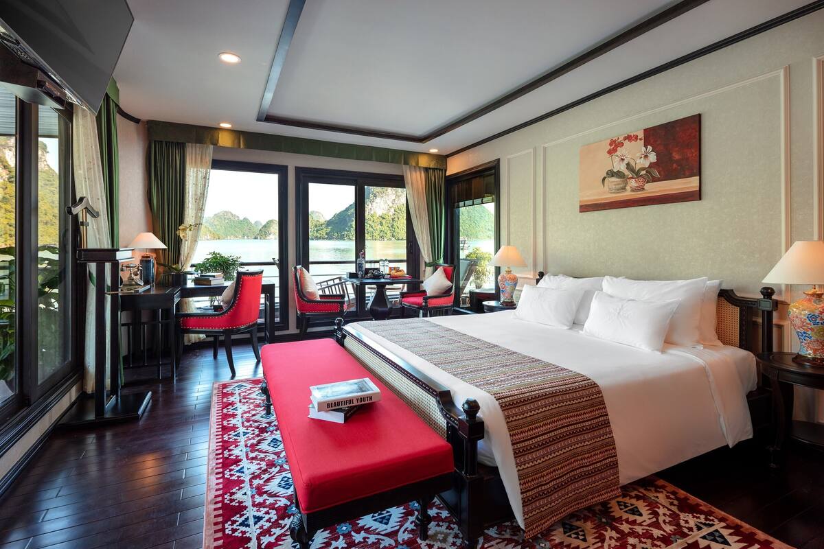 Quarto de hotel com uma cama grande, uma escrivaninha com cadeira, um banco e vista para o exterior.