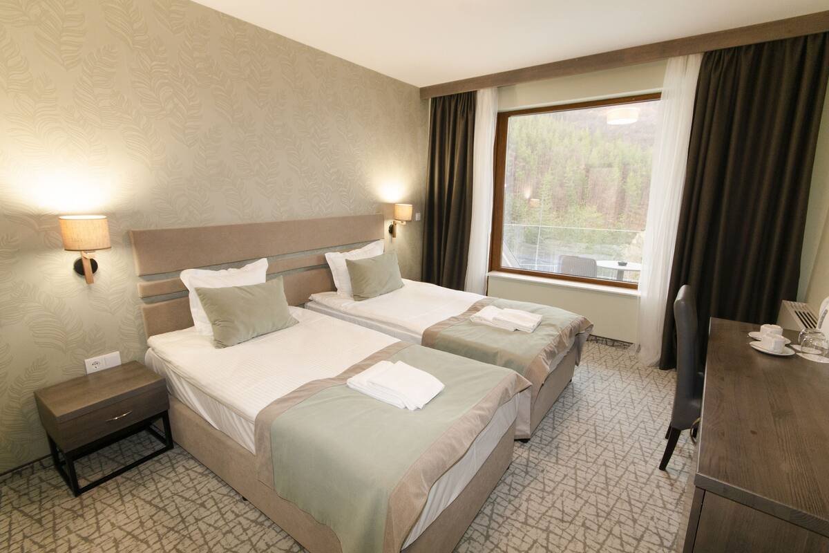 Quarto de hotel com duas camas, uma mesa de cabeceira, uma escrivaninha com cadeira e uma janela com cortinas.