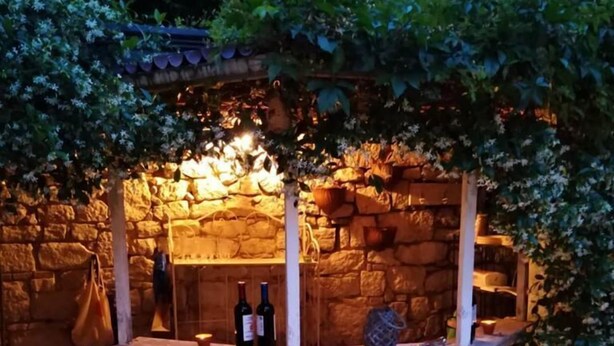 Uma área de bar ao ar livre com parede de pedra ao fundo, divisória de treliça branca e disposição de assentos com bancos acolchoados e cadeiras de metal.