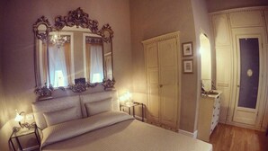 Quarto de hotel com uma cama grande, duas mesas de cabeceira, um guarda-roupa e um espelho com moldura decorada.