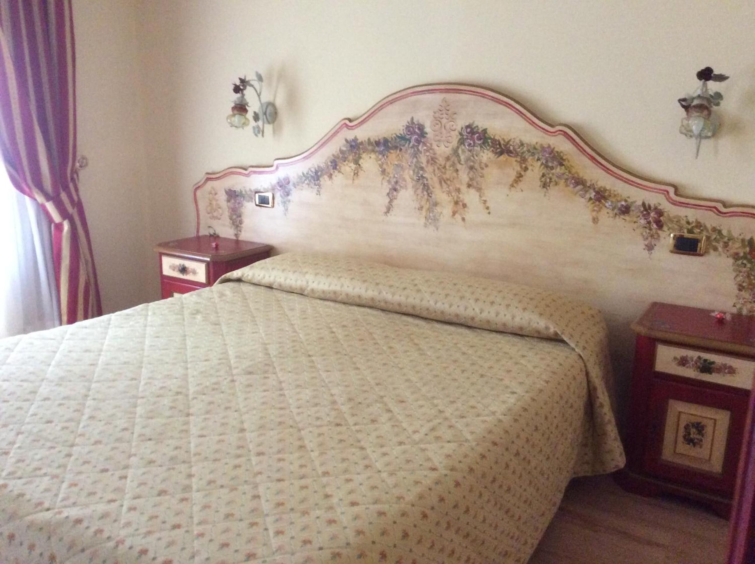 Cama bem arrumada com colcha estampada em florais, cabeceira de madeira com detalhes florais e duas mesas de cabeceira com pequenos objetos decorativos.