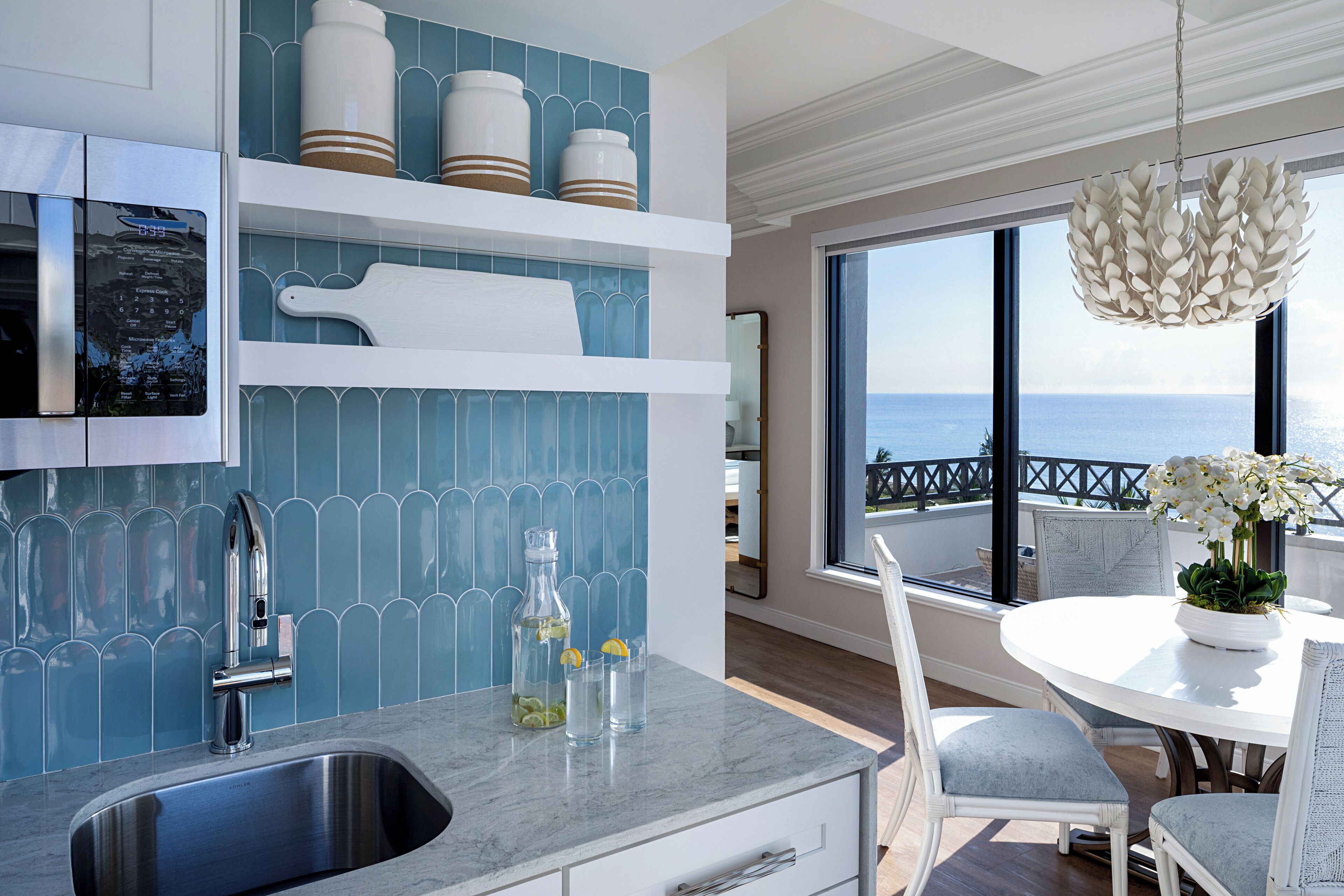 Cozinha moderna com pia de aço inoxidável, forno embutido e backsplash com azulejos azuis. Há uma área de jantar com mesa e cadeiras brancas, e uma grande janela com vista para o oceano.