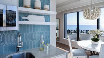 Cozinha moderna com pia de aço inoxidável, forno embutido e backsplash com azulejos azuis. Há uma área de jantar com mesa e cadeiras brancas, e uma grande janela com vista para o oceano.