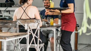 Ambiente de restaurante com mesa, cadeiras e um garçom servindo uma bebida.
