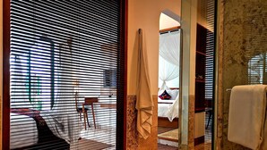 Quarto de hotel com uma cama, uma cadeira e uma mesinha. Há uma janela com persianas, uma toalha pendurada na parede e um espelho.