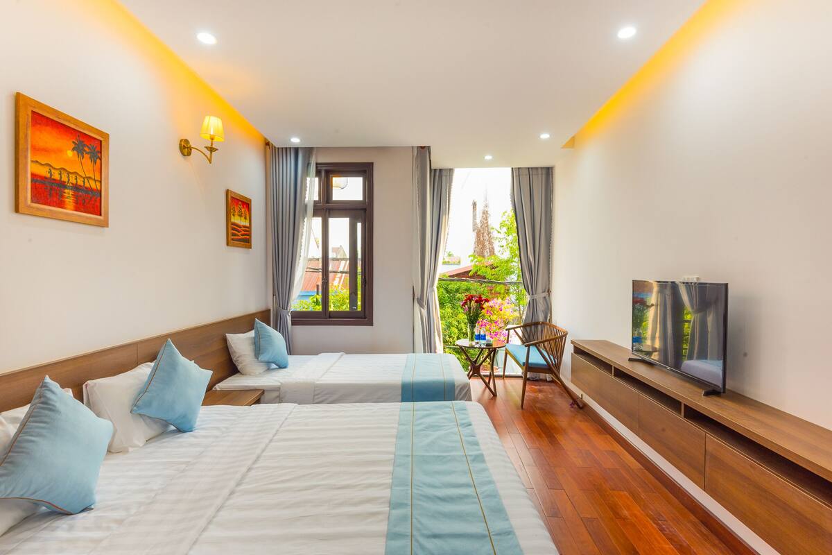 Quarto de hotel com duas camas, televisão e varanda com vista para área verde e um edifício.