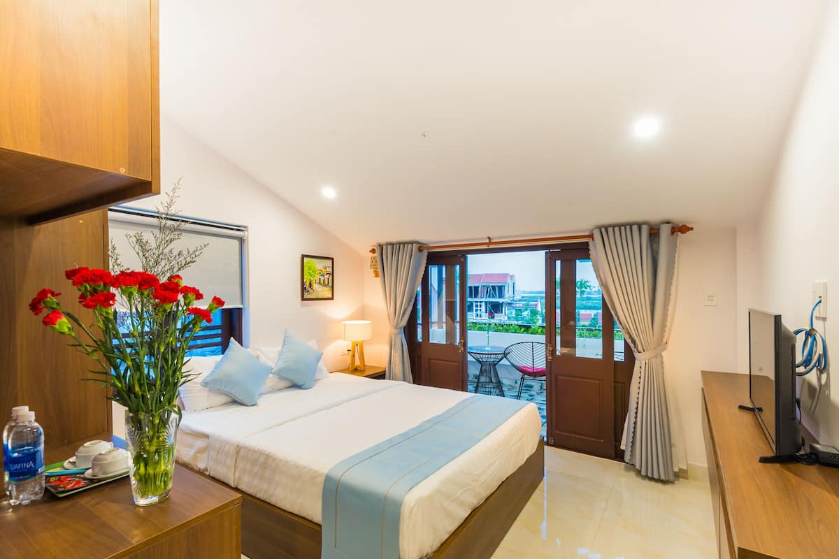 Quarto de hotel com cama, escrivaninha, televisão, janela com cortinas e um vaso com flores.
