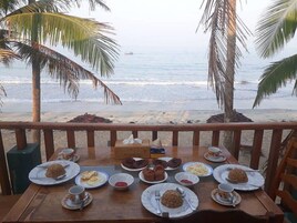 Mesa de madeira posta com pratos de comida e xícaras, com vista para uma praia com palmeiras e o mar.