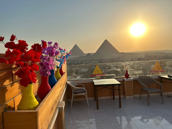 Um terraço com arranjos florais coloridos, uma mesa, cadeiras e vista para as pirâmides ao pôr do sol.
