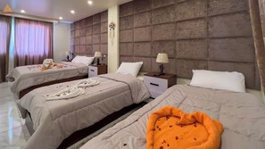 Quarto de hotel com três camas, cada uma com edredom branco e pétalas de rosa. Há mesas de cabeceira com abajures e um sonho apanhador na parede.