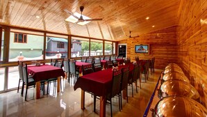 Restaurante com interior em madeira, amplas janelas e várias mesas cobertas por toalhas vermelhas.
