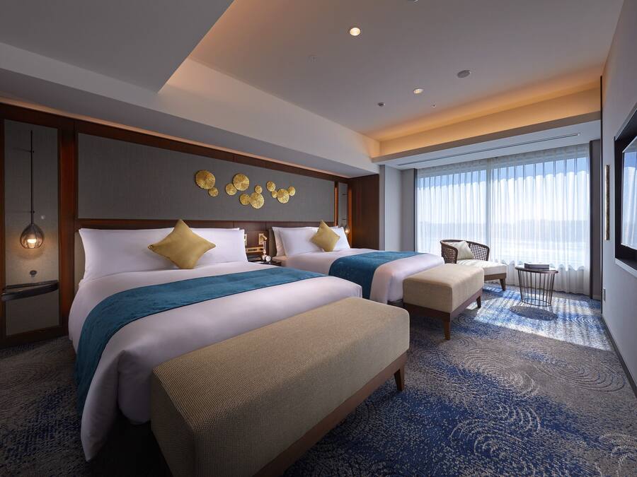 Quarto de hotel com duas camas, área de estar e uma janela grande com cortinas.