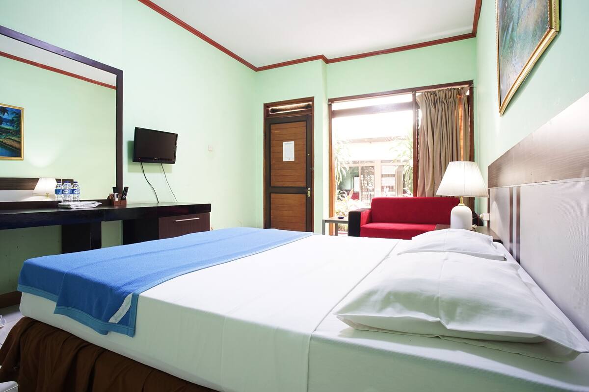 Quarto de hotel com cama, escrivaninha, televisão, uma cadeira vermelha e janela com cortinas.