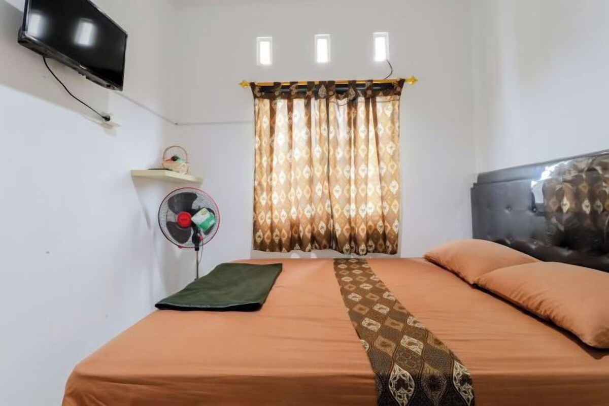 Quarto com cama, televisão montada na parede, ventoinha na mesa de cabeceira e janela com cortinas estampadas.