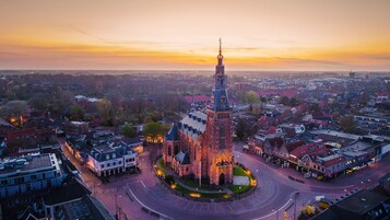 Uma igreja histórica com uma torre alta, rodeada por uma praça da cidade com edifícios e um cruzamento rotatório.