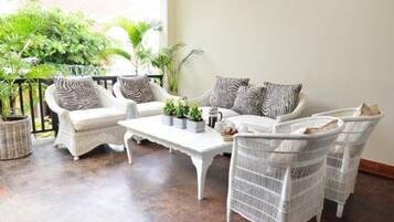 Um espaço de estar ao ar livre aconchegante, com móveis de vime e uma mesa branca, rodeado por vegetação exuberante.