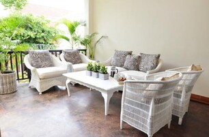Um espaço de estar ao ar livre aconchegante, com móveis de vime e uma mesa branca, rodeado por vegetação exuberante.