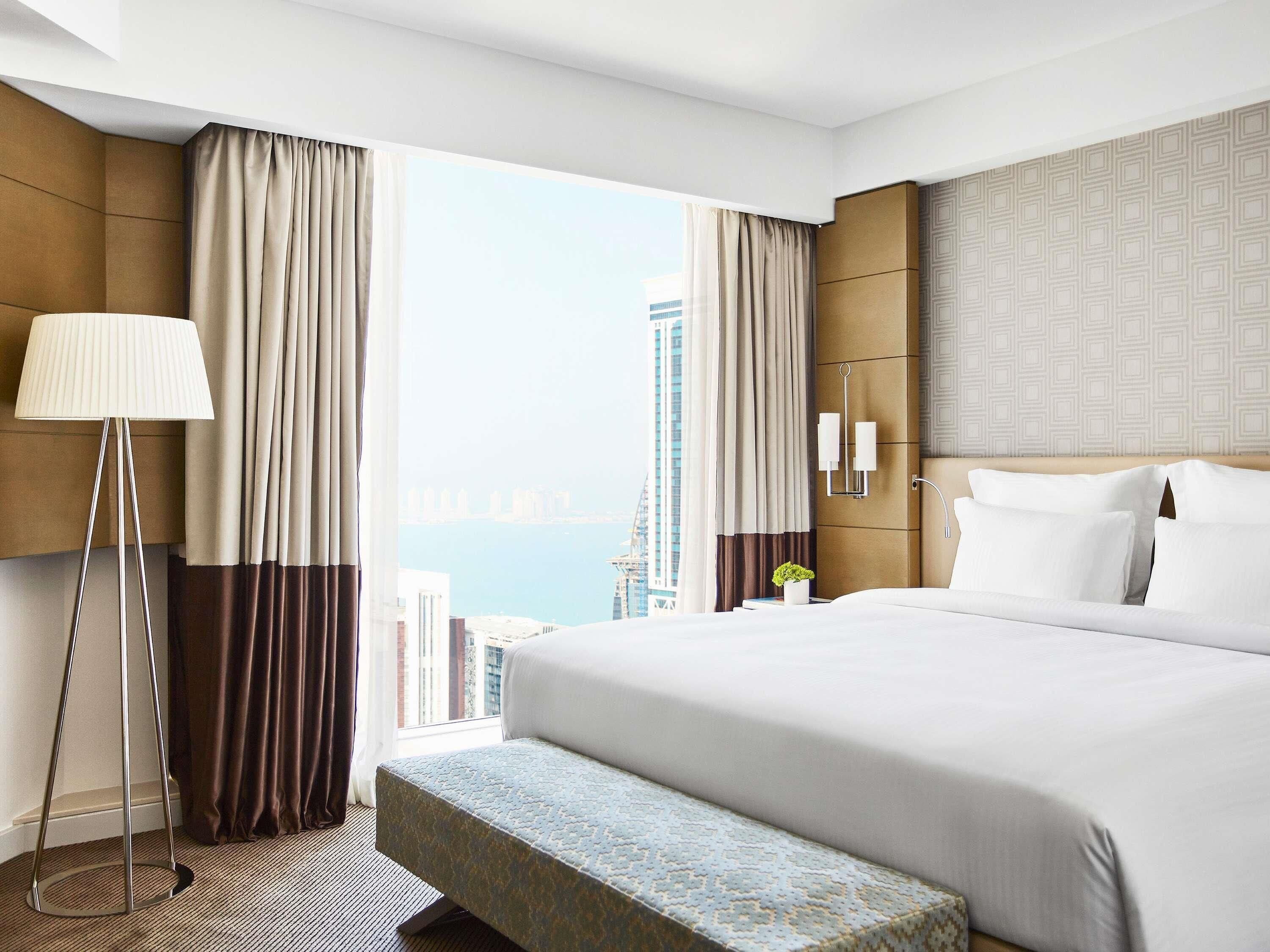 Quarto de hotel moderno com uma cama grande, uma mesinha de cabeceira, uma luminária e vista para a cidade.