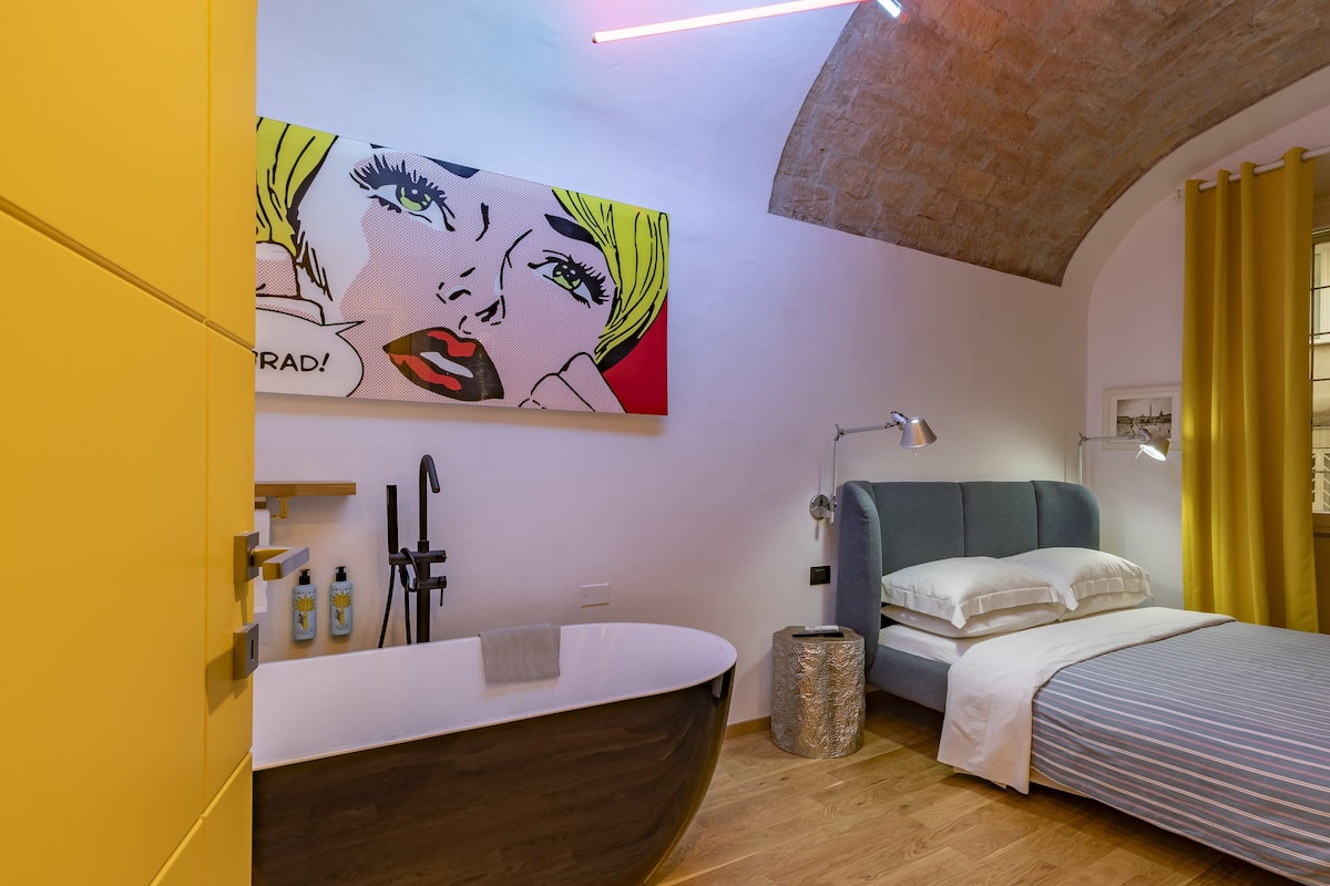 Um banheiro moderno com uma banheira independente, chuveiro e pia. Há uma cama com cabeceira azul e um abajur. Uma grande pintura pop art está na parede acima da cama.